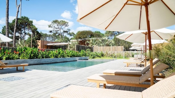 Área de piscina com espregui çadeiras e guarda-sóis, cercada por vegetação e um deck de madeira.
