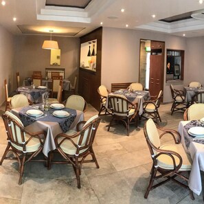 Uma sala de jantar com várias mesas e cadeiras, um lustre e uma televisão fixada na parede.