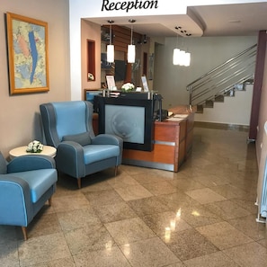 Uma área de recepção com balcão, dois poltronas azuis, uma mesa redonda pequena com uma flor e uma escadaria ao fundo.