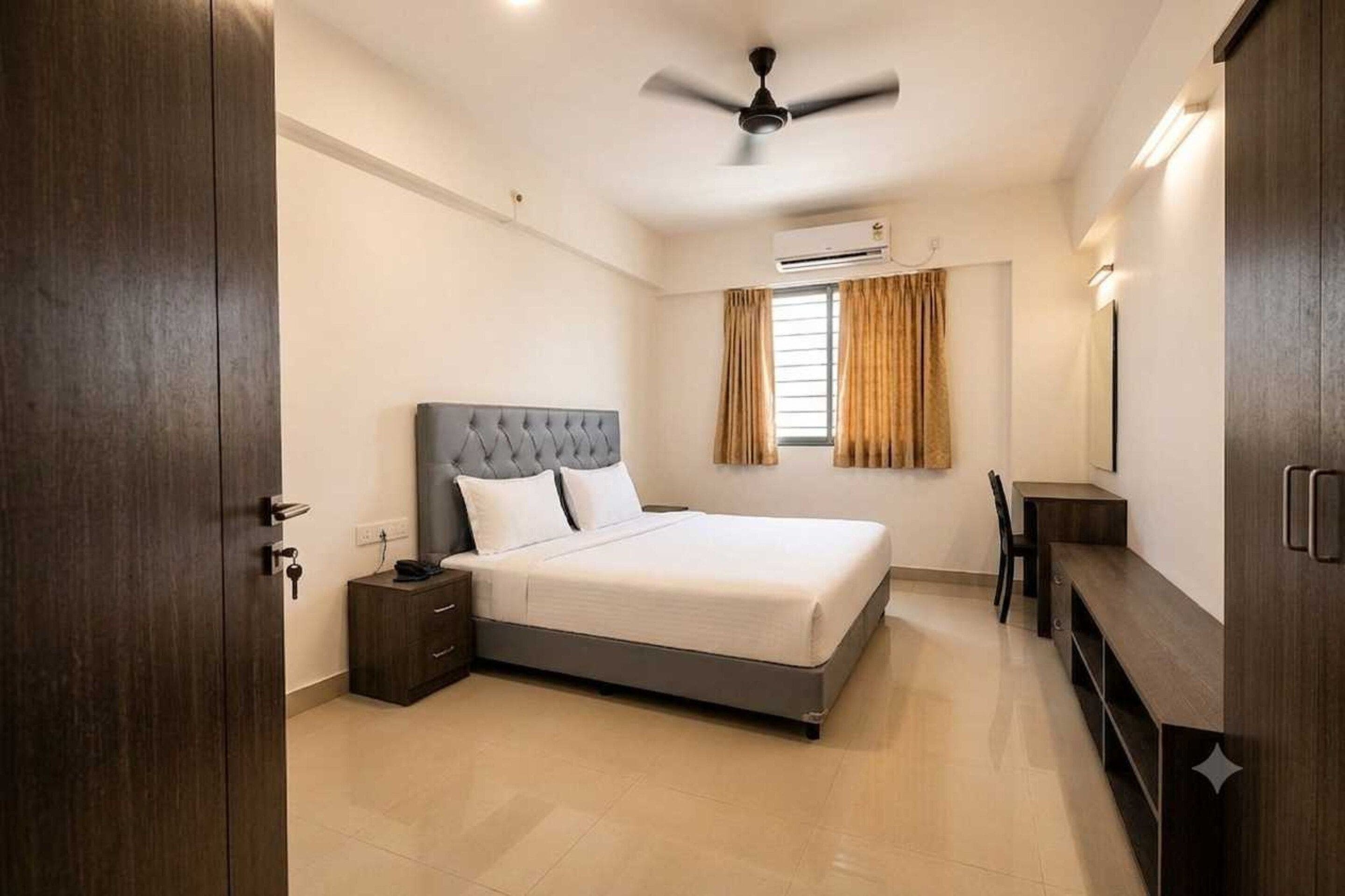 Quarto com cama, escrivaninha, cadeira, guarda-roupa, ventoinha no teto e ar condicionado.