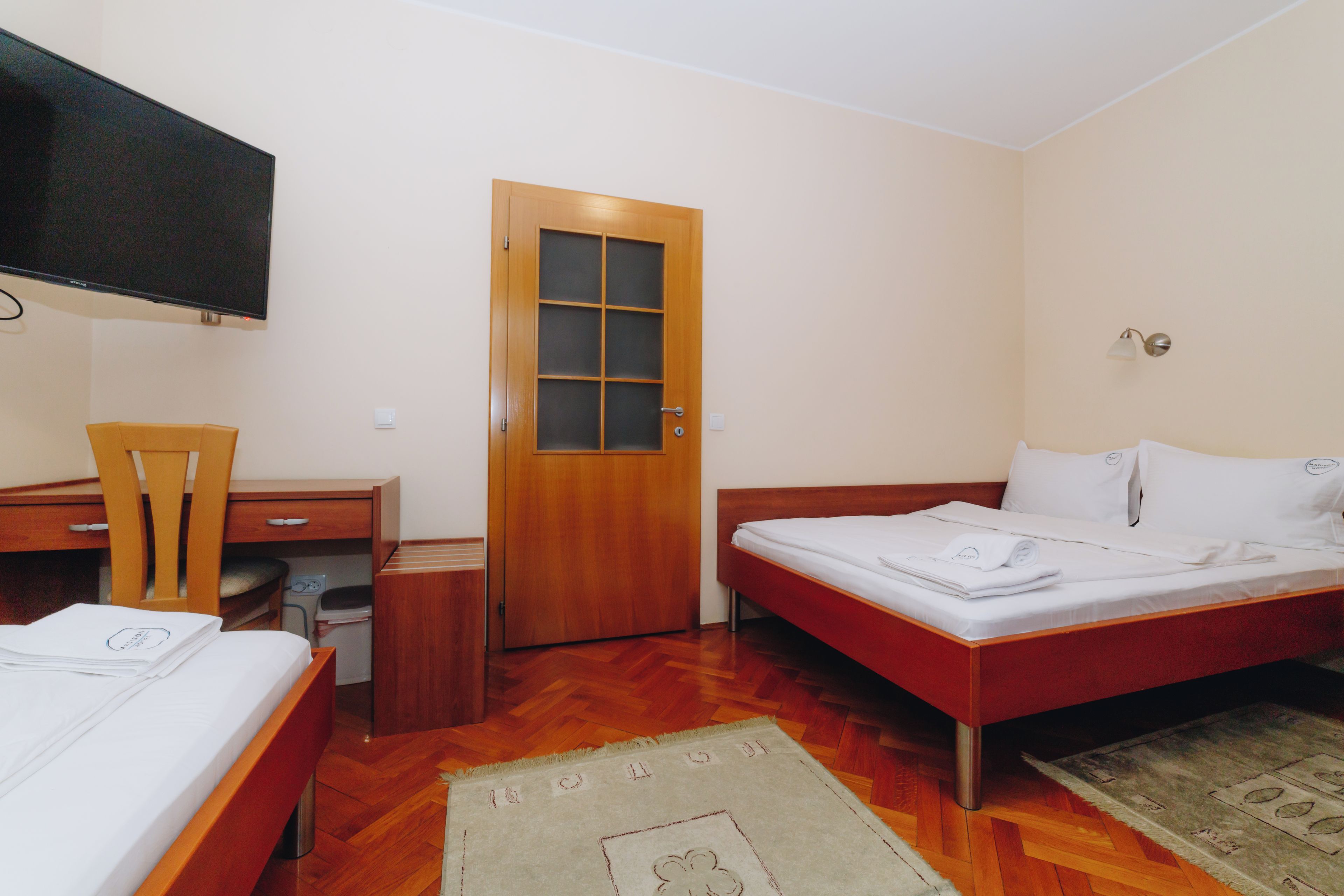 Quarto de hotel com cama, escrivaninha, cadeira, televisão e porta.