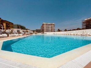 Piscina com cadeiras de descanso e um edifício ao fundo.
