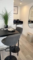 Espaço de jantar moderno com mesa posta para dois, uma planta em vaso e cozinha ao fundo.