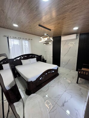 Quarto com cama de madeira, mesa de cabeceira, cadeira, espelho, luminária no teto, ar-condicionado fixado na parede e janela com cortinas.
