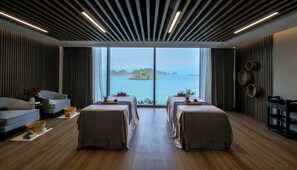Sala de spa com uma janela grande, duas macas de massagem, um sofá e piso de madeira.