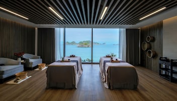 Sala de spa com uma janela grande, duas macas de massagem, um sofá e piso de madeira.