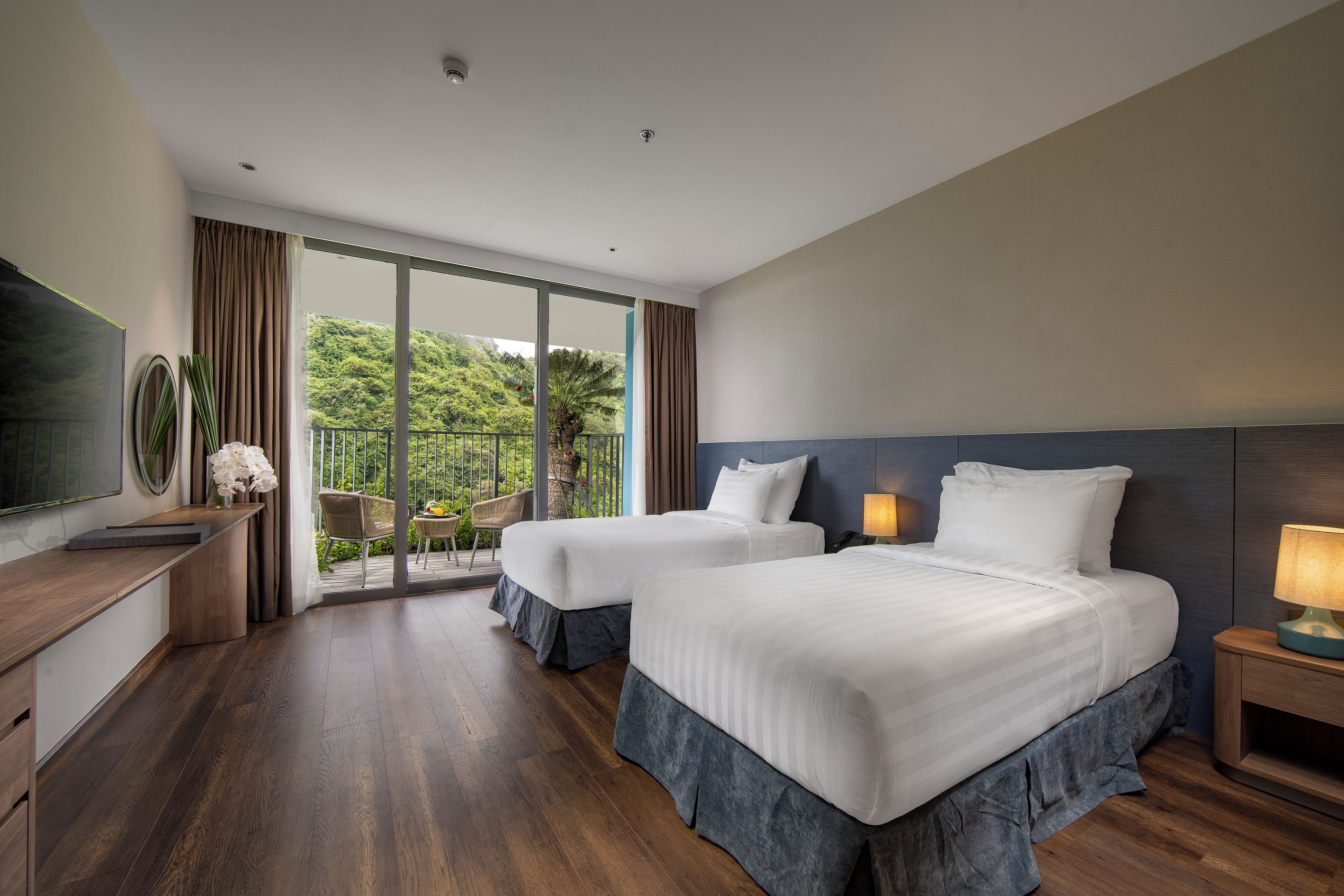 Quarto de hotel com duas camas, uma mesa de escritório, televisão e varanda com vista para áreas verdes.