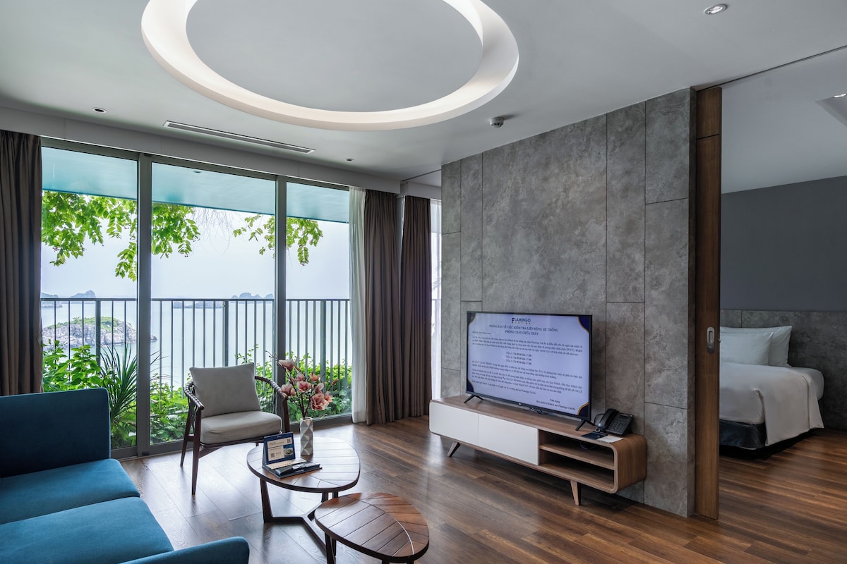 Quarto de hotel moderno com televisão grande, sofá, mesa de centro e varanda com vista para o mar.