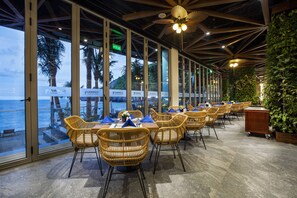 Restaurante com cadeiras e mesas de vime, amplas janelas com vista para o mar e um ventilador de teto.