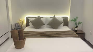 Cama bem arrumada com cabeceira, dois travesseiros, mesa de cabeceira com relógio e uma pequena planta.