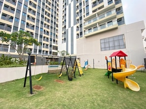 Um parquinho com balanços e escorregadores, cercado por uma área gramada e um prédio residencial alto ao fundo.