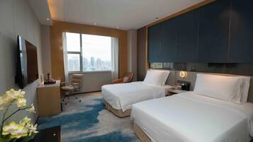 Quarto de hotel com duas camas, uma escrivaninha, uma cadeira, televisão e uma grande janela com vista para a cidade.