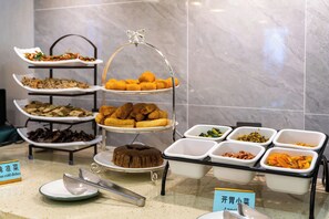 Buffet com diversos pratos, incluindo uma variedade de cozinha asiática, dispostos em suportes escalonados e em travessas.