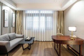 Quarto de hotel com sofá, mesa pequena, escrivaninha com luminária e janela com cortinas.