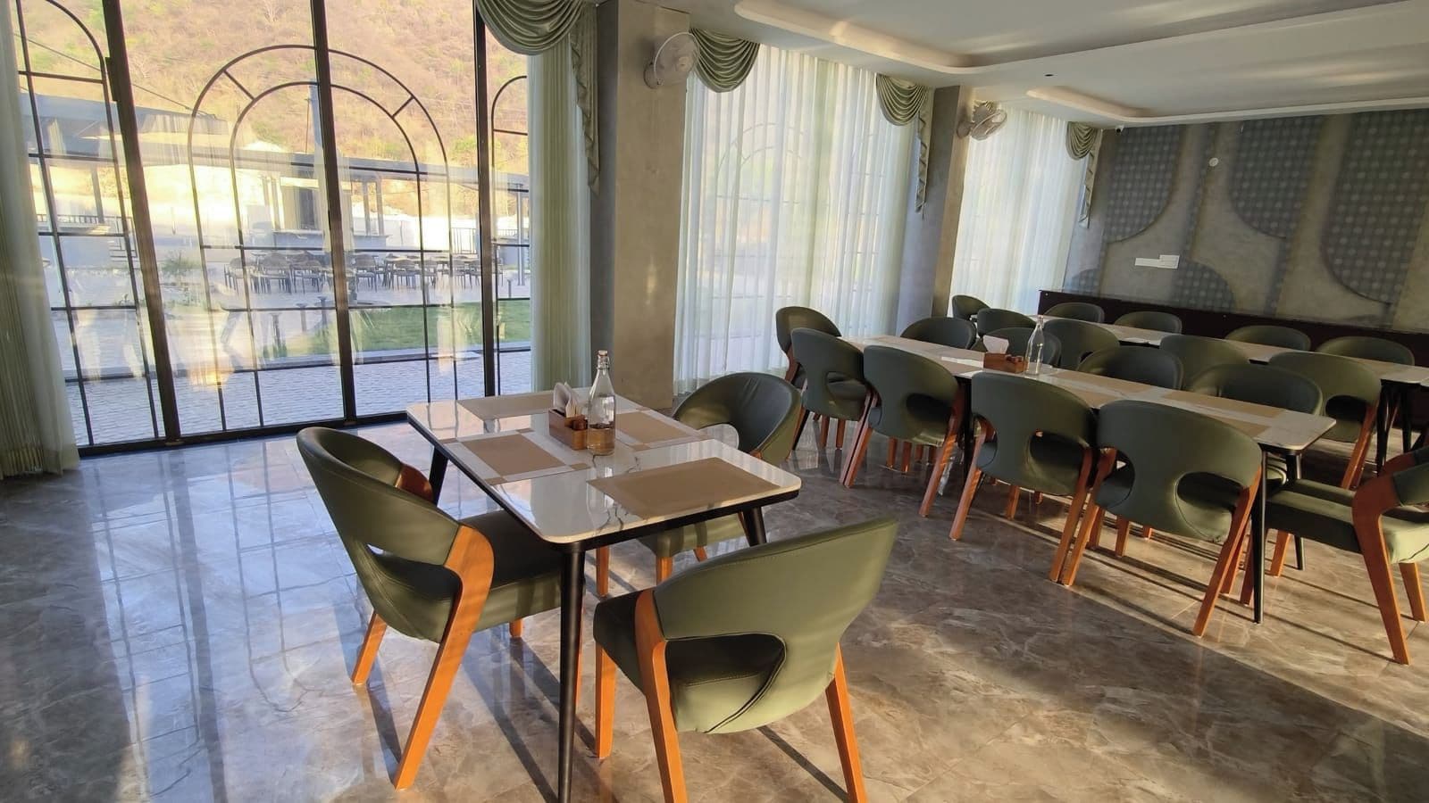 Uma área de refeições com mesas e cadeiras, grandes janelas com cortinas e vista para um corpo de água.