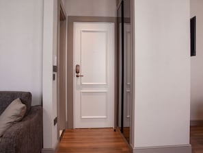 Um corredor com uma porta branca fechada, um sofá cinza com um almofado e piso de madeira.
