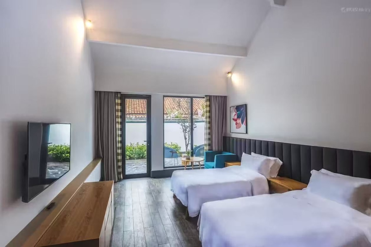 Quarto de hotel com televisão, duas camas, um sofá e vista pela porta de vidro.