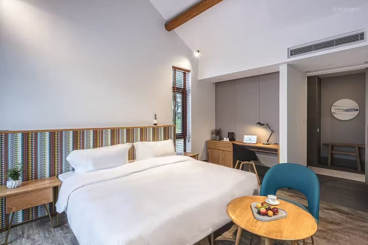 Quarto de hotel moderno com uma cama grande, uma escrivaninha, uma cadeira e uma mesinha redonda com frutas.