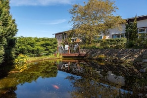 Uma casa com uma ponte de pedra sobre um lago, rodeada por vegetação exuberante e árvores.