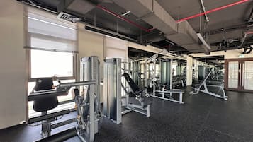 Academia com diversos equipamentos de exercício, amplas janelas e piso preto salpicado.