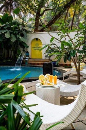 Área da piscina com placa para Staymaster Veridian, uma cadeira branca, uma mesa com bebida e uma pequena cascata.