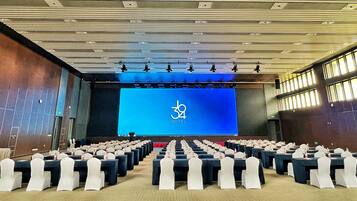 Uma sala de conferências com fileiras de mesas e cadeiras, uma tela grande exibindo o número 34 e uma área de palco.