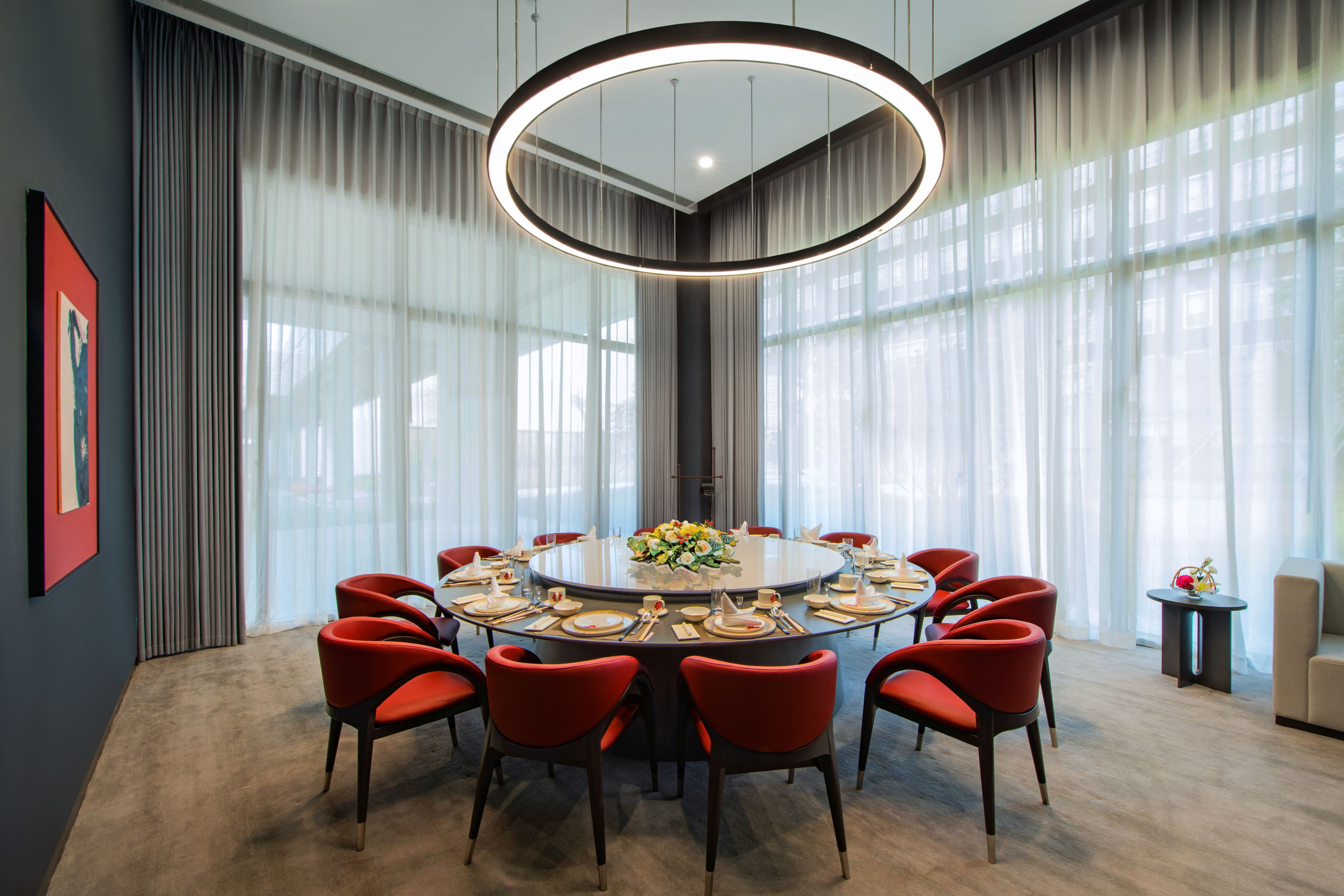 Uma sala de conferências moderna com uma mesa redonda, cadeiras vermelhas e um arranjo floral central.