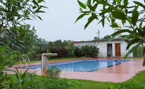Piscina com plataforma de mergulho, rodeada por vegetação e um edifício com telhado vermelho.