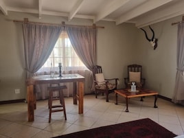 Uma área de refeições com mesa de madeira, duas cadeiras, um banco e uma mesinha de centro. Há uma janela com cortinas, um enfeite de chifres na parede e piso de azulejos.