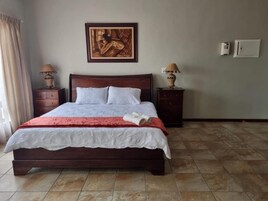 Cama com colcha branca e vermelha, duas mesas de cabeceira com abajures, ar-condicionado fixado na parede e uma moldura com imagem sobre a cama.