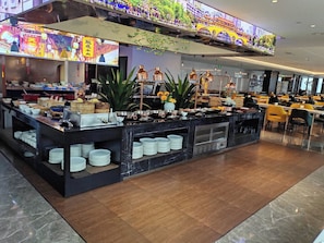 Uma área de buffet com diversos pontos de serviço, uma variedade de pratos e uma grande tela digital exibindo conteúdo colorido.