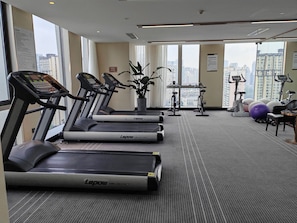 Academia com esteiras, bicicletas ergométricas e bolas de exercício, com vista para a cidade através de amplas janelas.