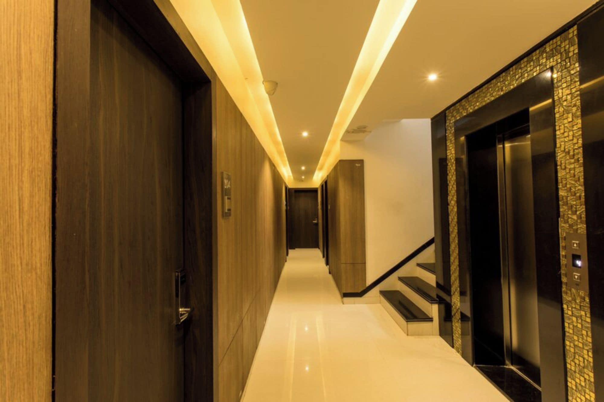 Um corredor moderno de hotel com portas de madeira, uma escadaria e um elevador.