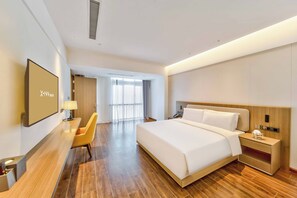 Quarto de hotel com uma cama grande, uma escrivaninha com cadeira, televisão e mesa de cabeceira com abajur.