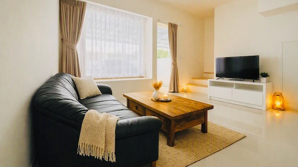Uma sala de estar moderna com um sofá de couro preto, uma mesa de centro de madeira, uma TV de tela plana sobre um suporte branco e uma grande janela com cortinas.