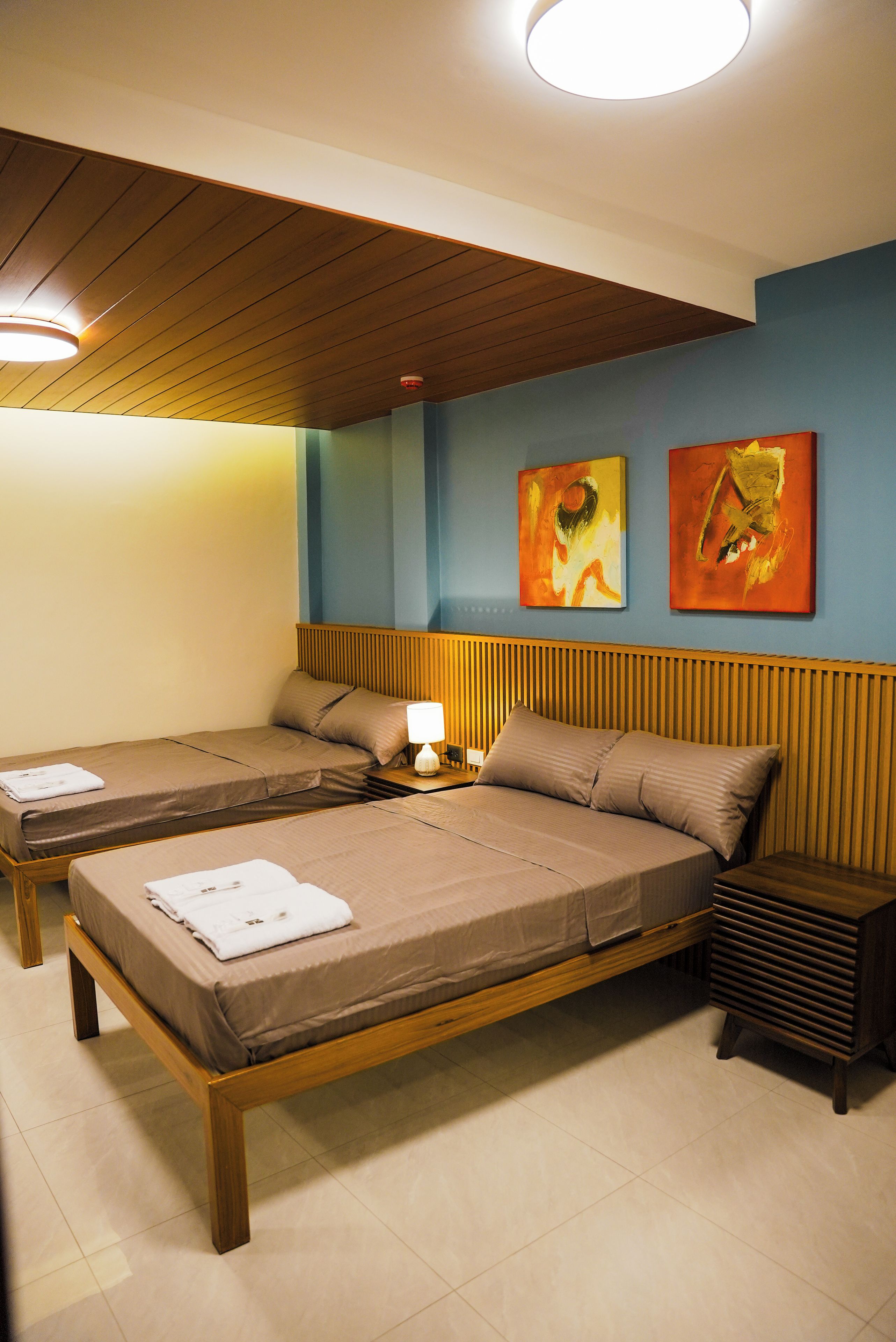Quarto de hotel com duas camas, uma mesinha de cabeceira, um abajur e duas pinturas na parede.