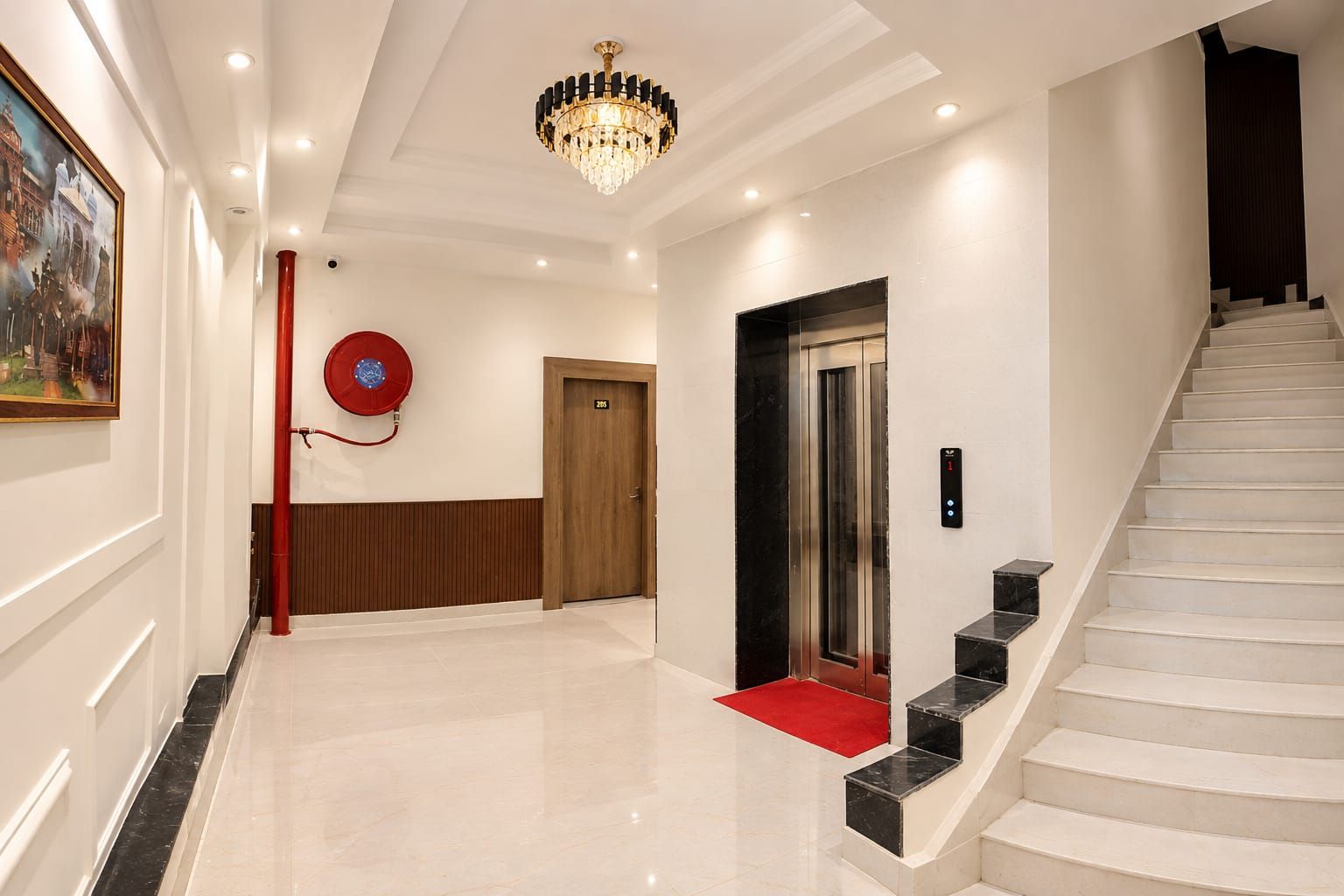 Um hall de hotel moderno com piso de mármore, um candelabro, um extintor de incêndio vermelho, uma porta de madeira, um elevador e uma escadaria.