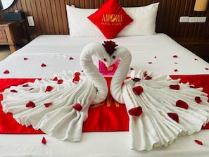 Quarto de hotel com cama, travesseiro vermelho com a inscrição 'AMORII HOTEL & CAFE' e arranjo de toalhas em formato de cisnes com pétalas de rosa.