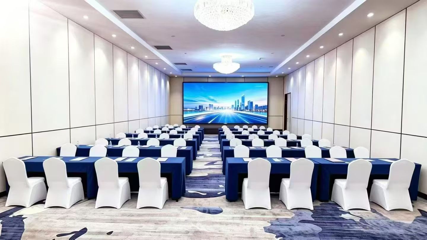 Uma sala de conferências com fileiras de mesas e cadeiras, uma tela grande na frente e um candelabro.