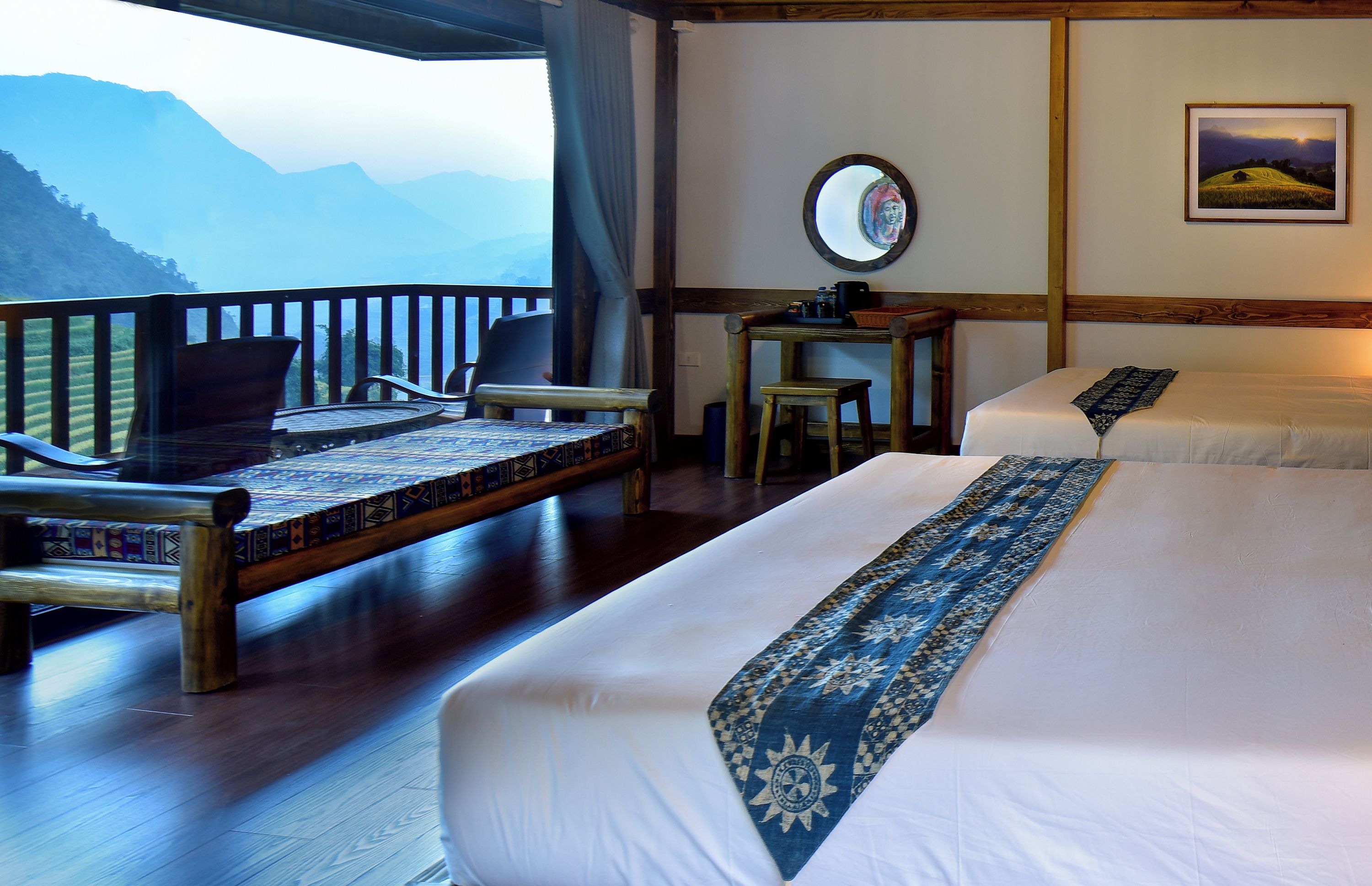 Quarto de hotel com varanda, uma cama com colcha estampada, uma mesa pequena com cadeira e um espelho fixado na parede.