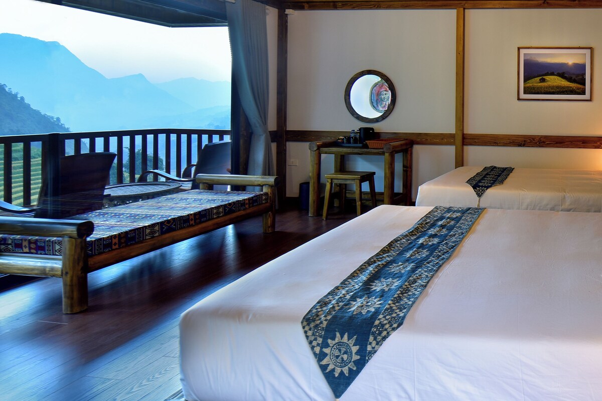 Quarto de hotel com varanda, uma cama com colcha estampada, uma mesa pequena com cadeira e um espelho fixado na parede.