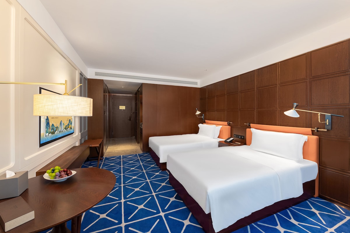 Quarto de hotel com duas camas, uma escrivaninha com luminária, televisão e um tapete azul estampado.