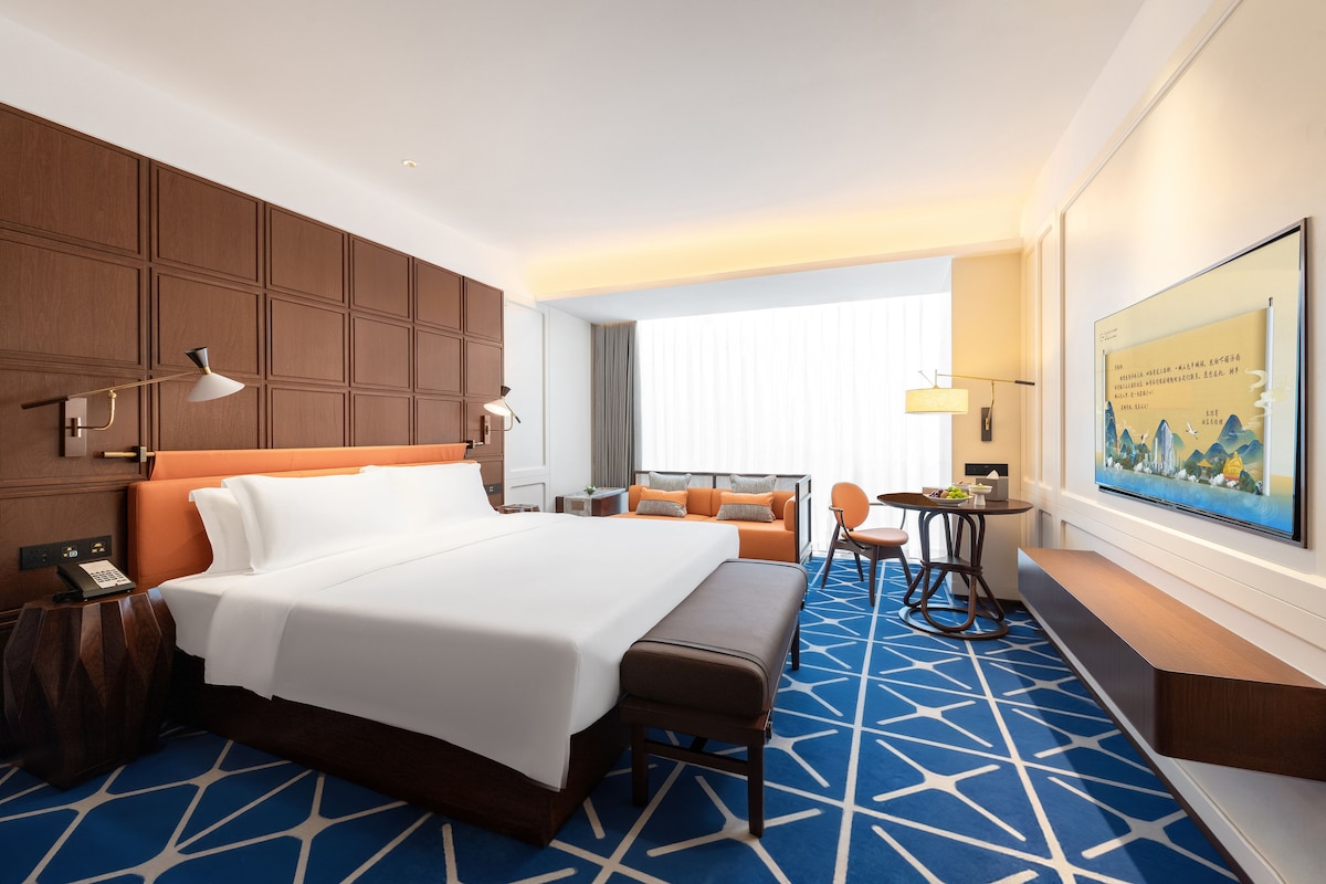 Quarto de hotel com uma cama grande, área de estar com uma mesa e um quadro na parede.