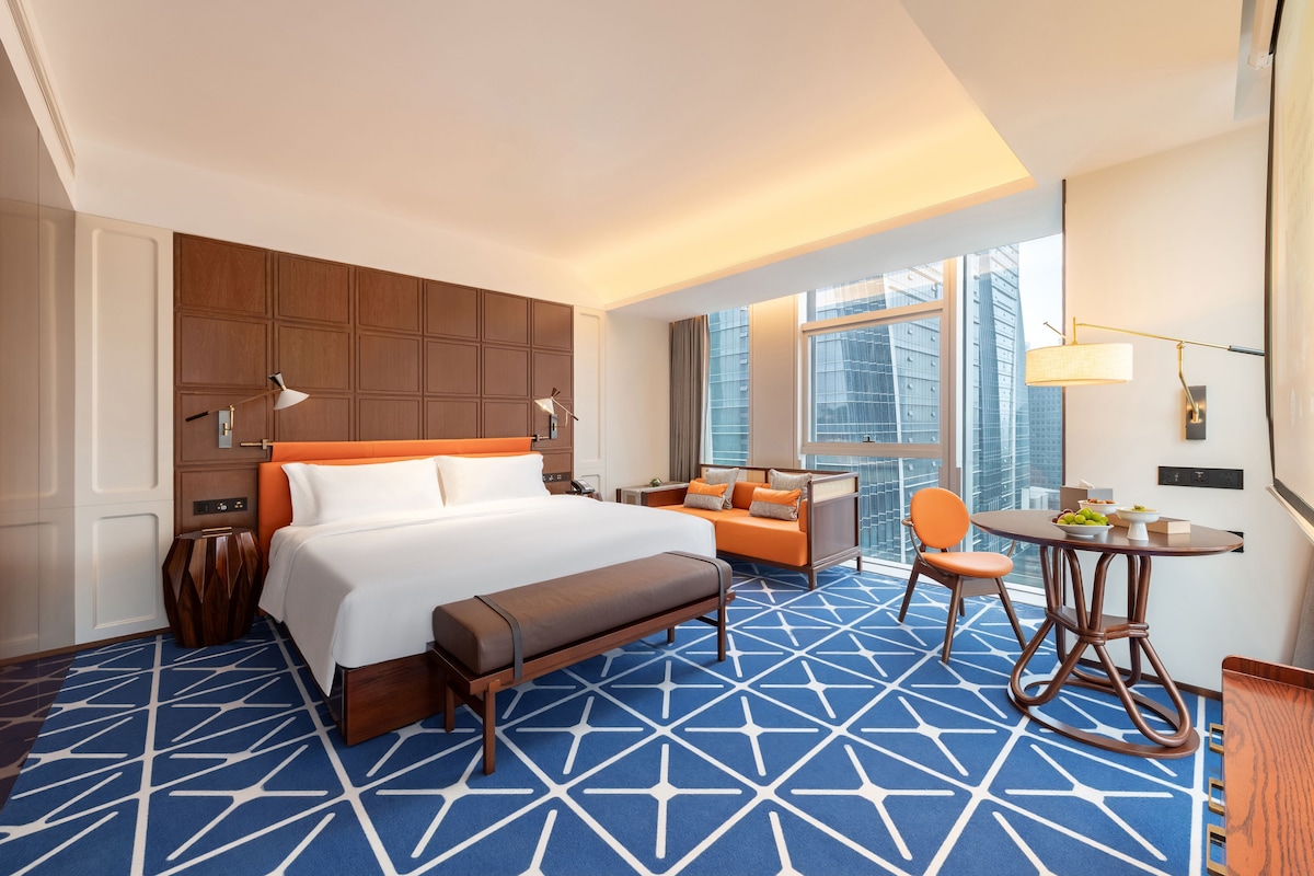 Quarto de hotel com uma cama grande, espaço para sentar, uma mesinha e vista para a paisagem urbana.