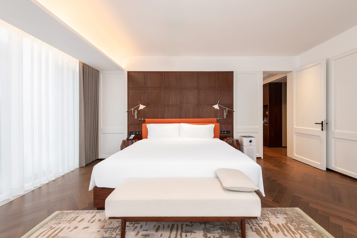 Quarto de hotel com uma cama grande, mesas de cabeceira, luminárias e uma janela com cortinas.