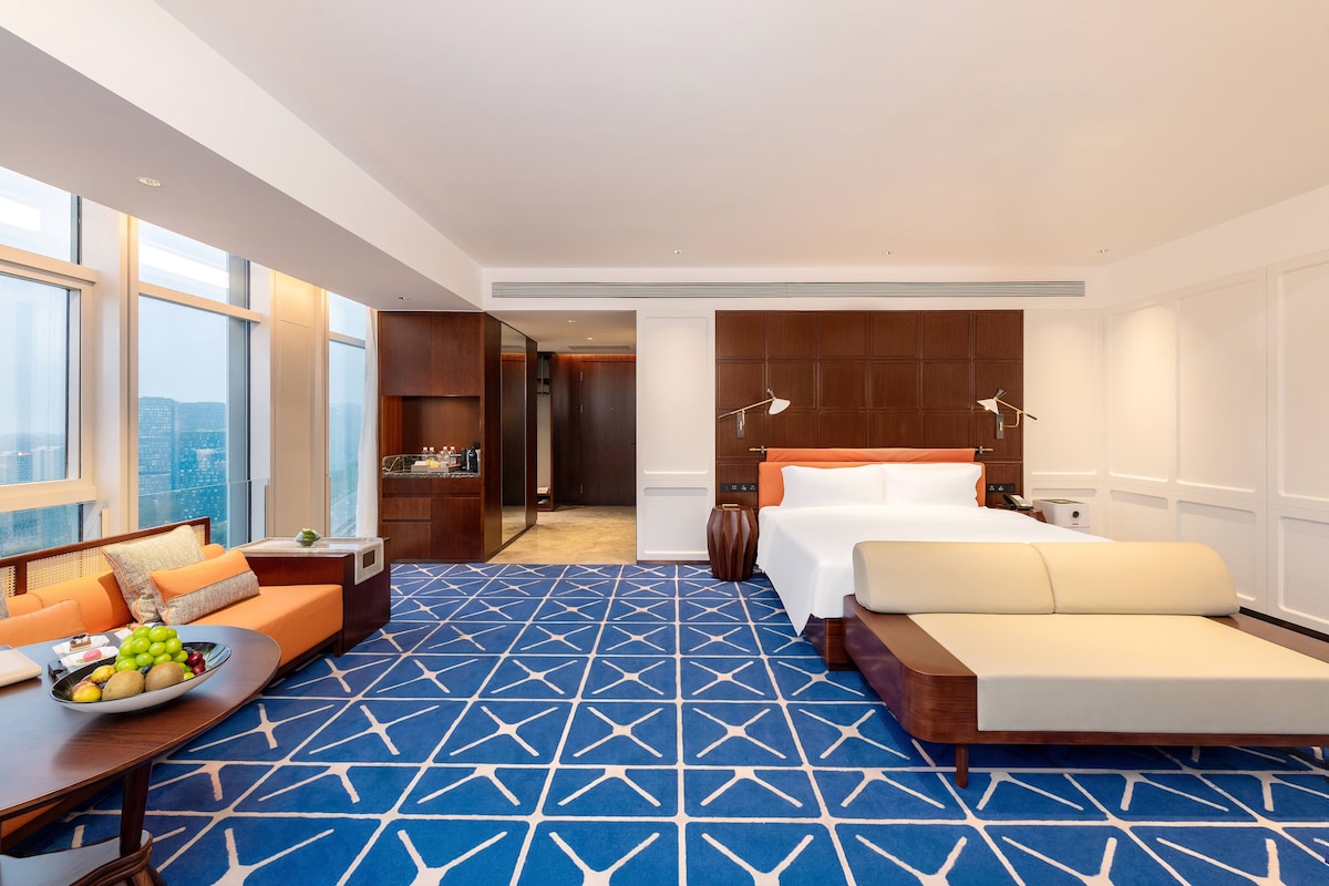 Quarto de hotel com cama, sofá, mesa de jantar com frutas, uma janela grande e vista para a cidade.