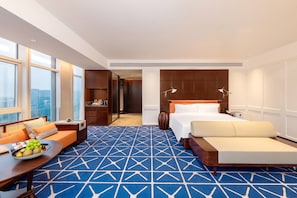 Quarto de hotel com cama, sofá, mesa de jantar com frutas, uma janela grande e vista para a cidade.