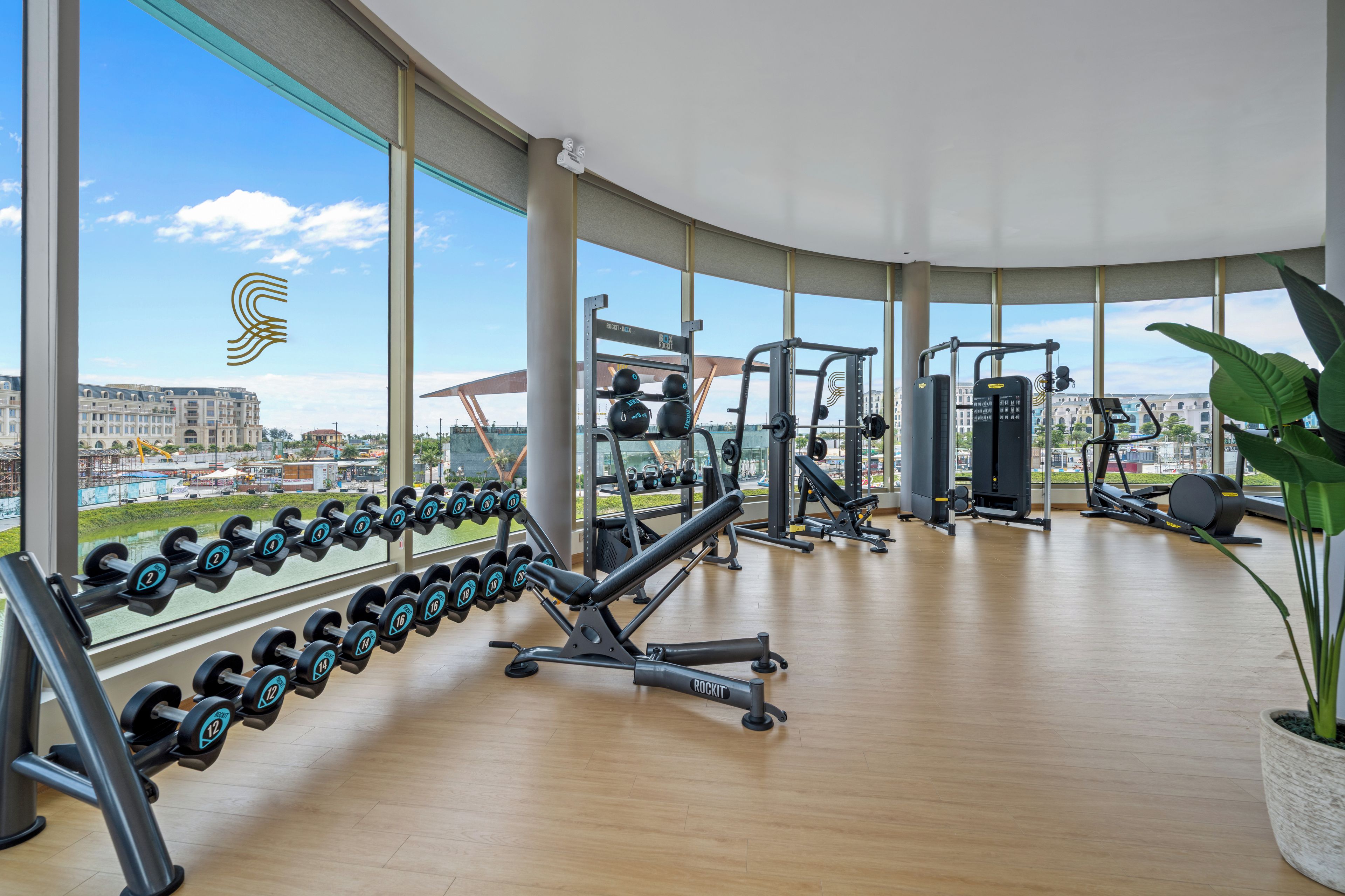 Uma academia com equipamentos de musculação, uma máquina de remo e uma janela ampla com vista para edifícios e um canteiro de obras.