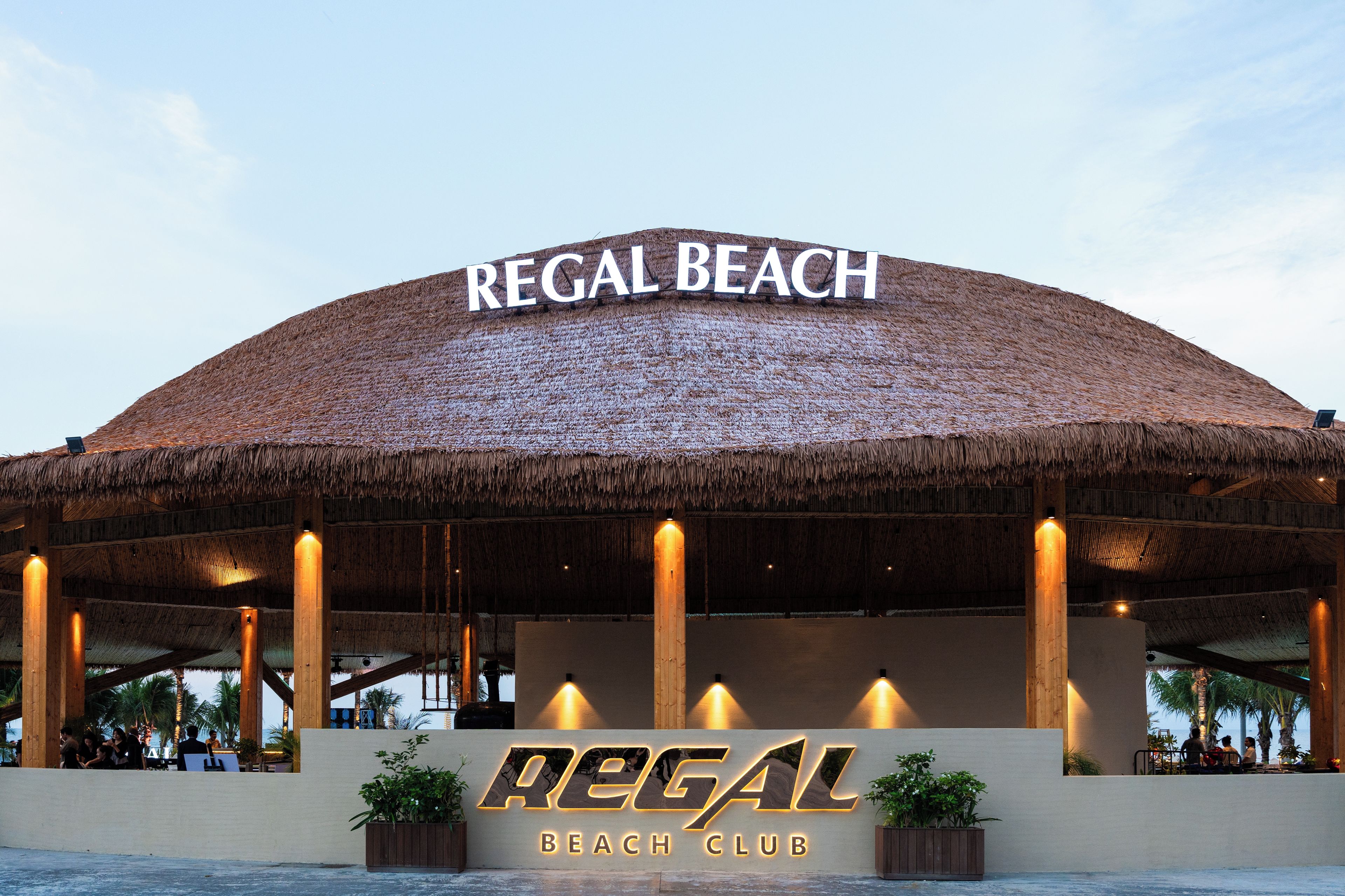 Um edifício com telhado de palha e a frase 'Regal Beach Club' iluminada na fachada.