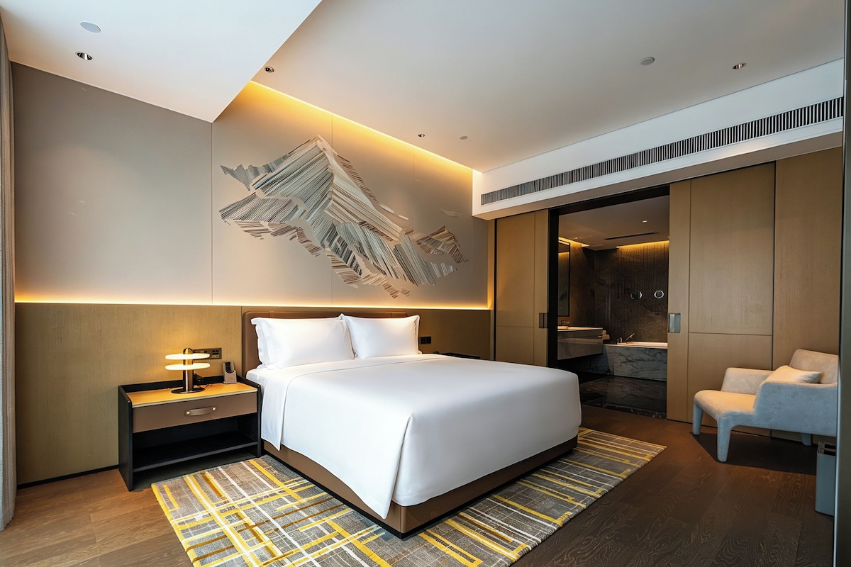 Quarto de hotel moderno com uma cama grande, uma mesa de cabeceira, uma escrivaninha, uma cadeira e um banheiro visível através de uma porta aberta.